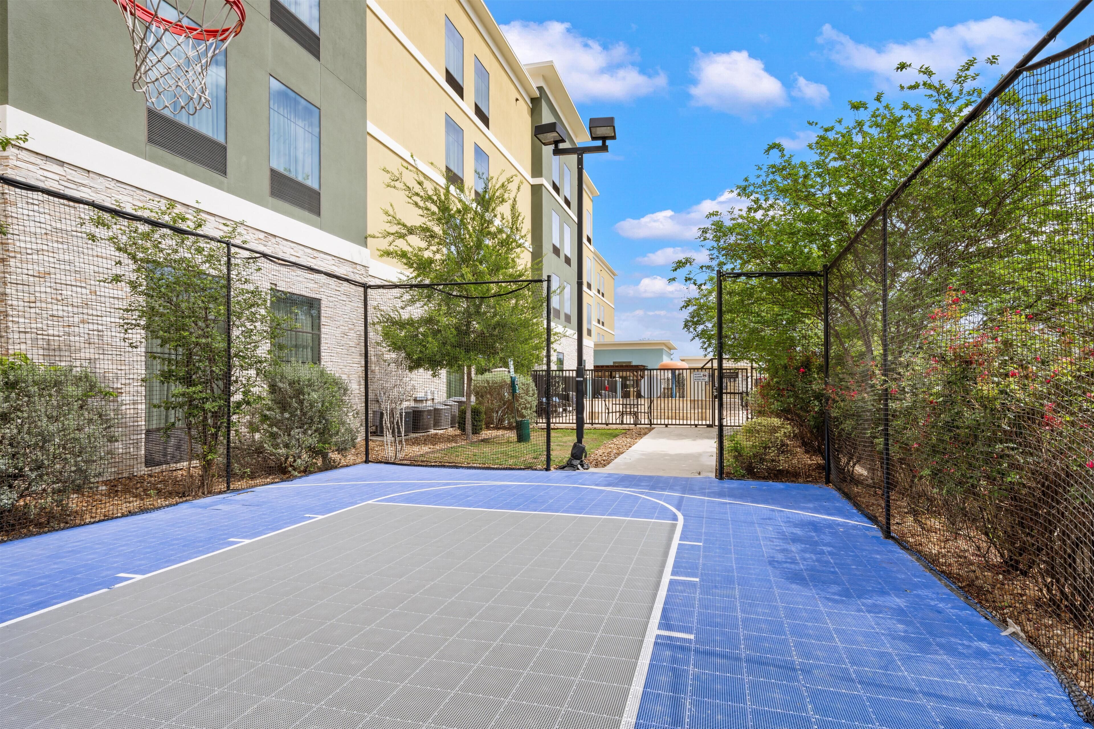 Uma quadra de basquete ao ar livre com superfície azul, cercada por uma grade, árvores e um edifício com janelas.