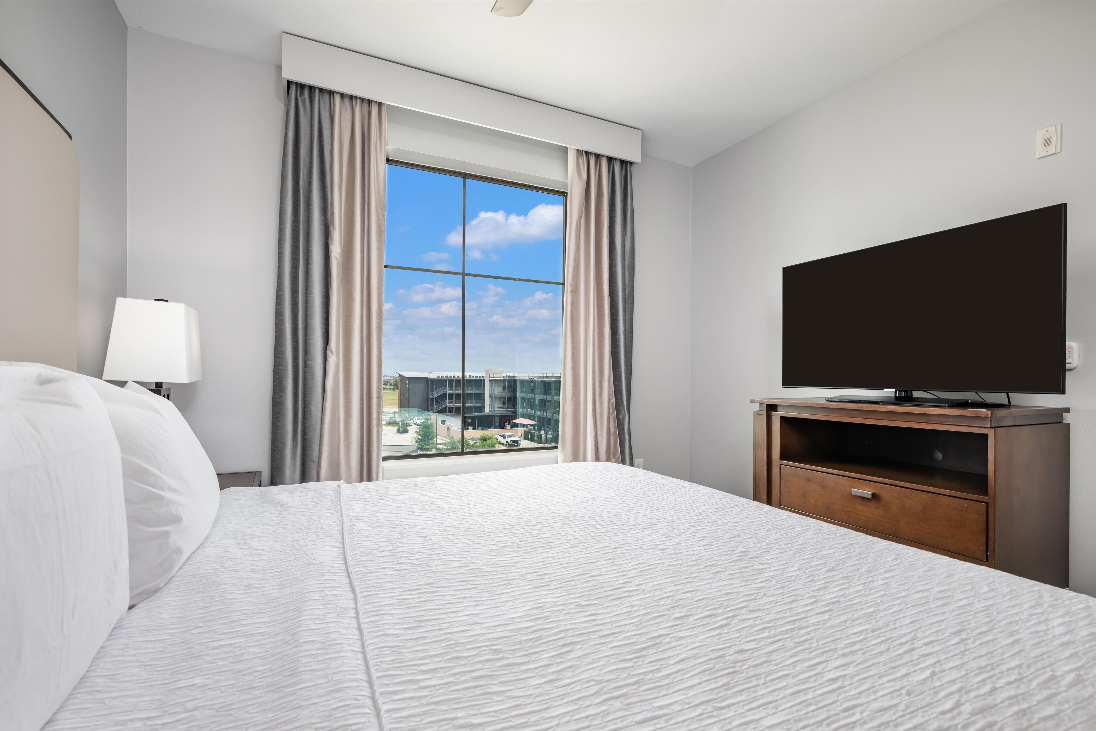 Quarto de hotel com uma cama grande, televisão, janela com cortinas e vista para edifícios.