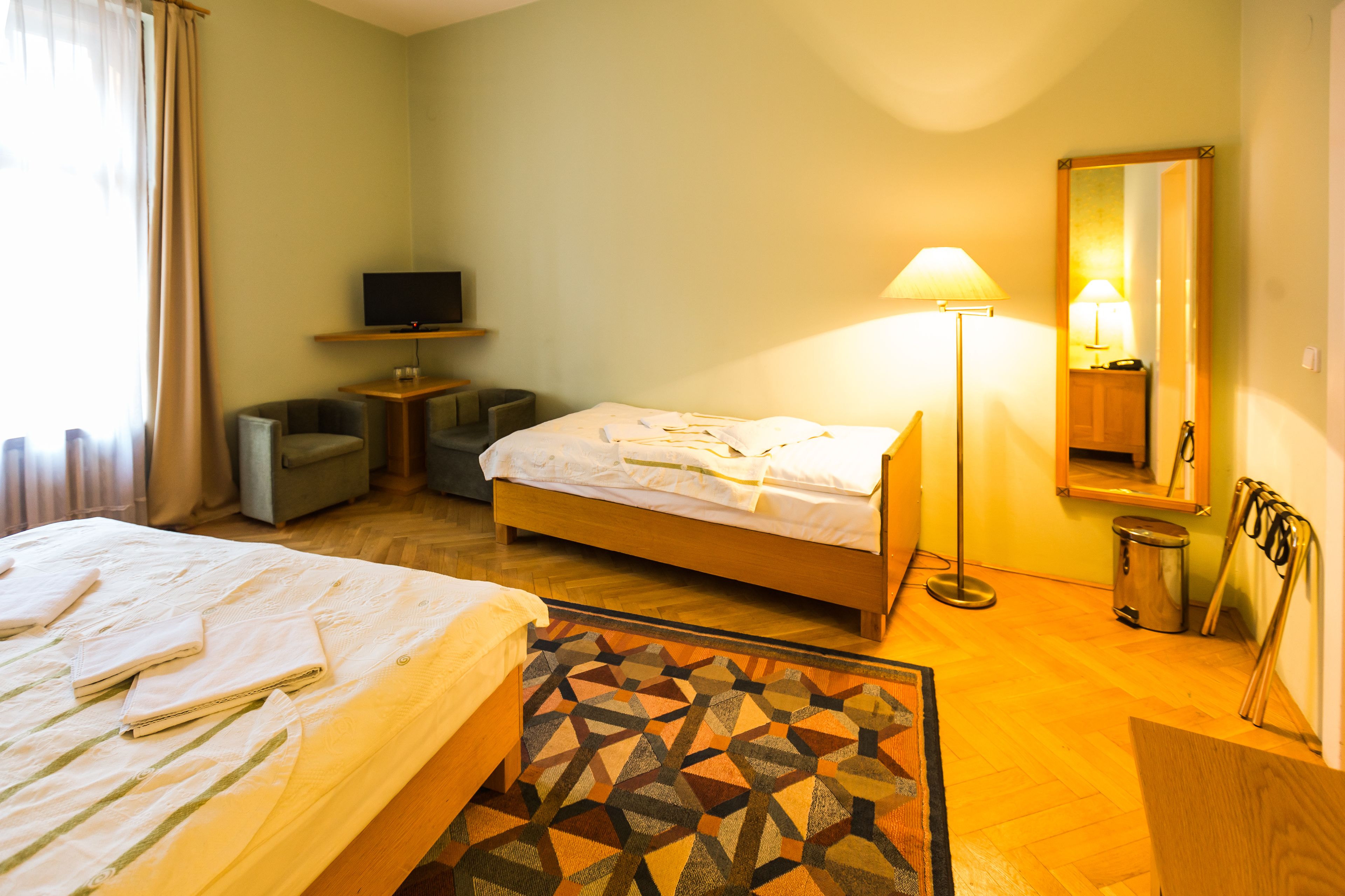 Quarto de hotel com cama, escrivaninha, cadeira, televisão, abajur, espelho e tapete estampado.