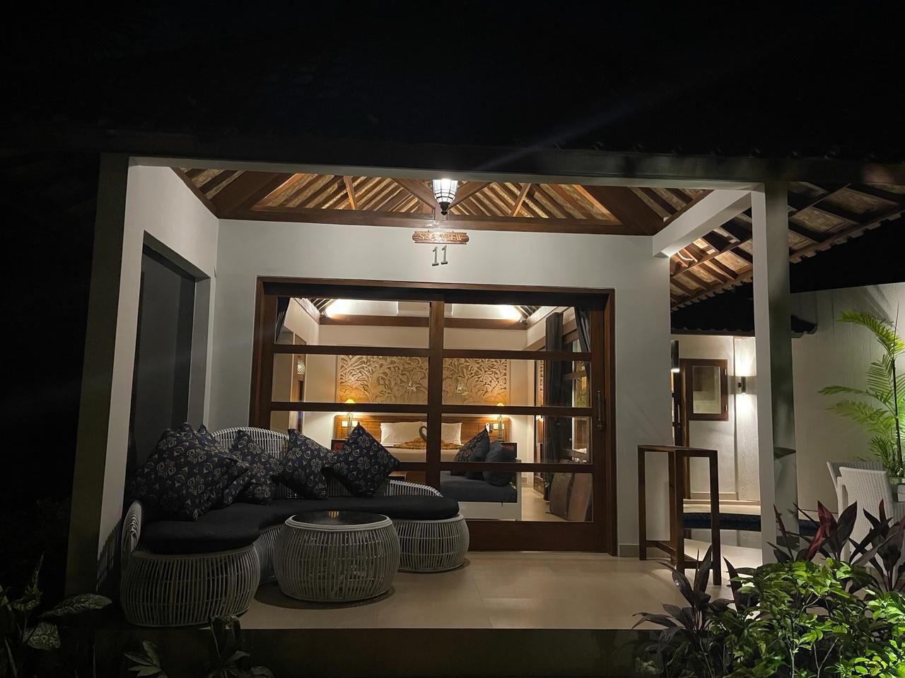 Uma área de estar moderna ao ar livre, com sofá e cadeiras, teto de madeira e vista para um quarto com cama e mesa de cabeceira.
