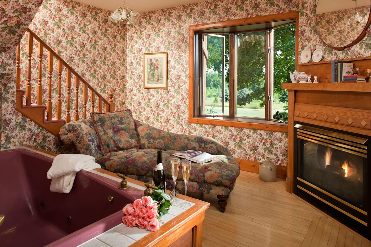 Quarto com papel de parede floral, escada de madeira, sofá, lareira e janela com vista para o verde.