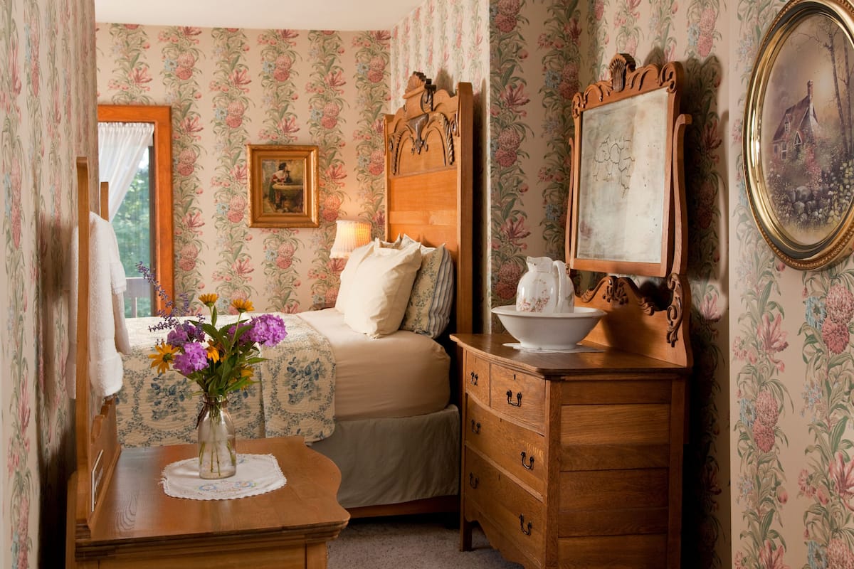 Quarto com papel de parede floral, cama de madeira, cômoda, espelho e um vaso com flores sobre uma mesa de cabeceira.