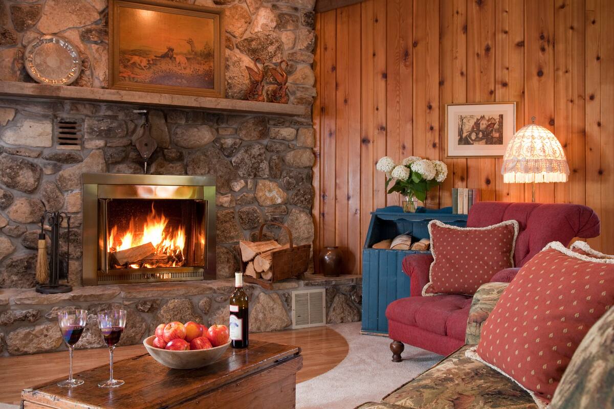 Uma sala de estar acolhedora com lareira de pedra, uma poltrona vermelha e uma mesa de centro de madeira com copos de vinho e uma tigela de frutas.