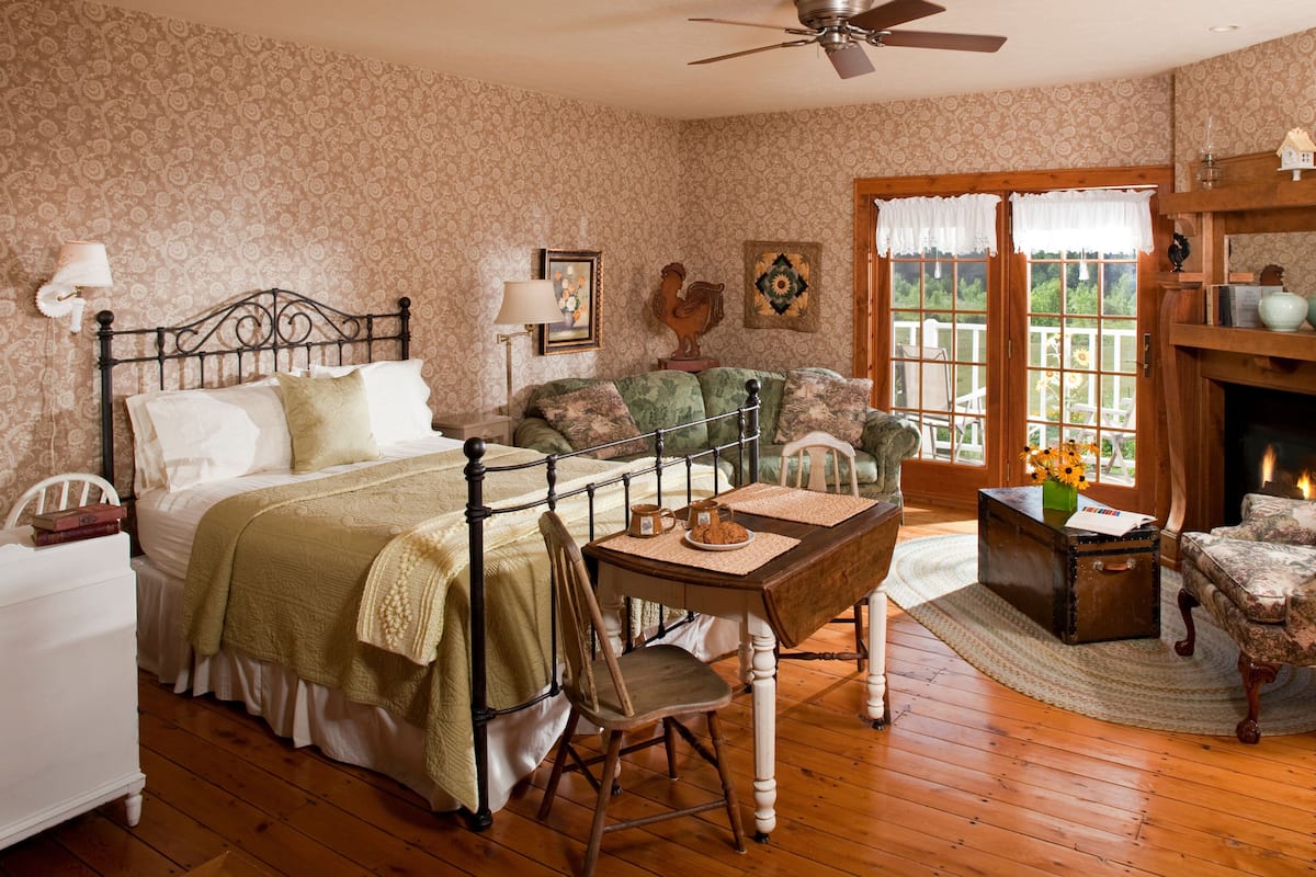 Quarto aconchegante com cama, uma cadeira de madeira, uma mesa com um vaso e uma lareira.