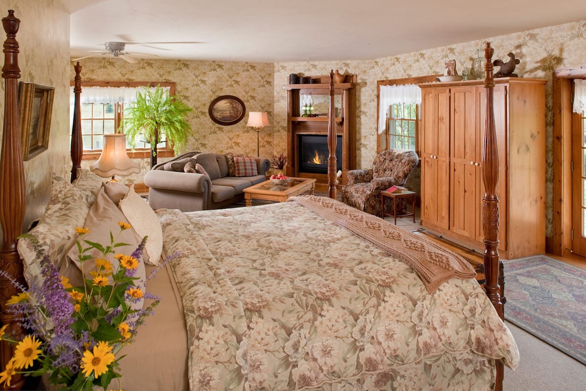 Quarto tradicional com uma cama grande, um espaço de estar com sofá e poltrona, um guarda-roupa de madeira e uma lareira.