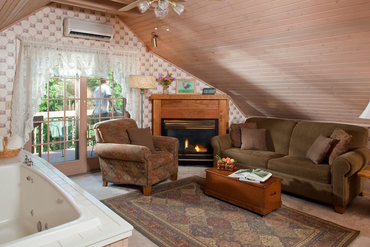 Uma sala de estar aconchegante com lareira, sofá, poltrona estampada e uma grande janela com vista para uma varanda.