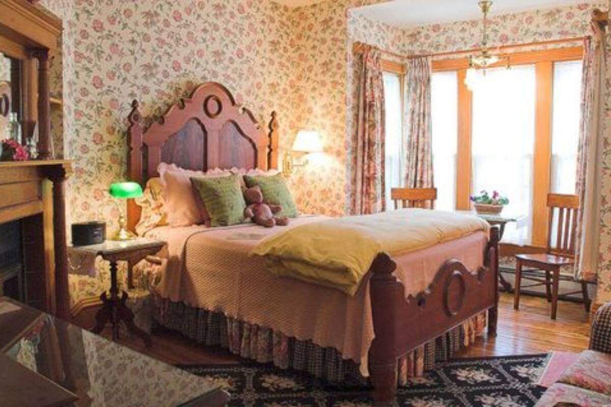 Quarto com papel de parede floral, cama de madeira com cabeceira, mesa de cabeceira com abajur e uma cadeira.