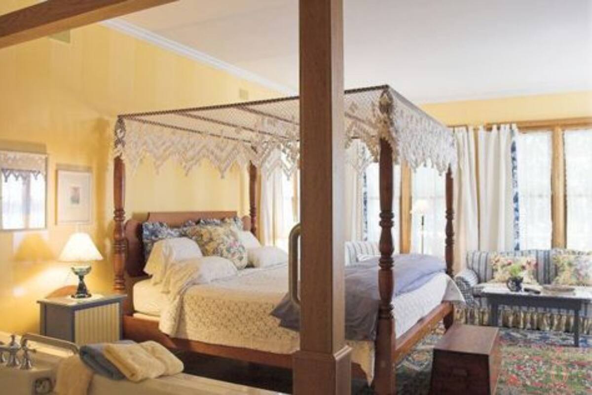 Uma cama de dossel com cortina, uma mesa de cabeceira com abajur, uma cadeira com almofada florida e uma pequena mesa com um vaso.