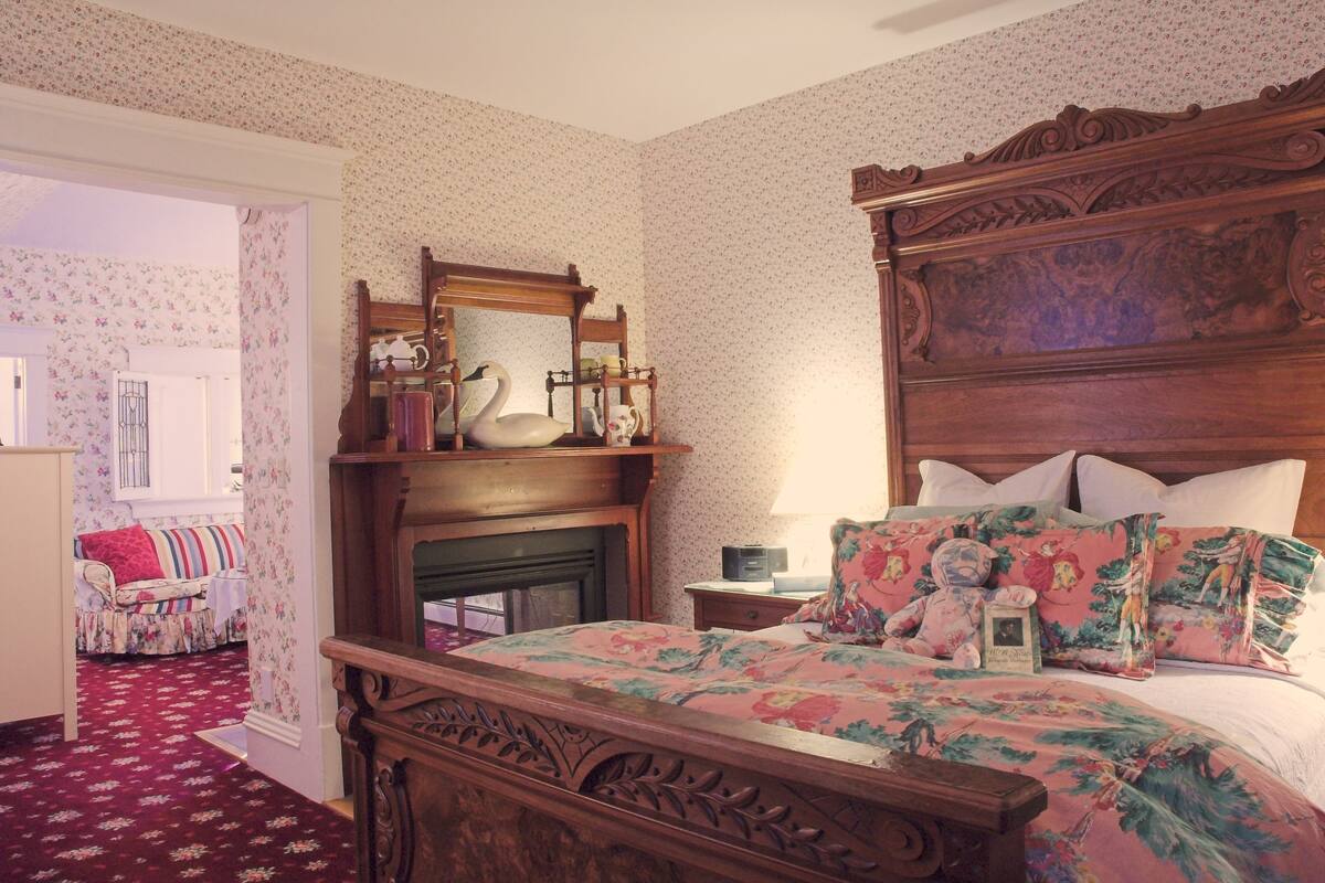 Quarto com cama de madeira, papel de parede floral, espelho sobre a cama, uma mesinha com abajur e uma cadeira com almofada floral.