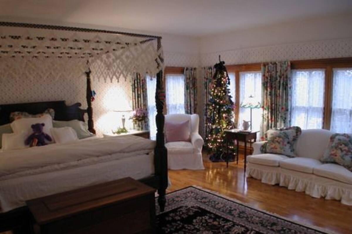 Um quarto com uma cama de dossel, mesa de cabeceira, banco de madeira, poltrona branca, sofá e uma árvore de Natal.