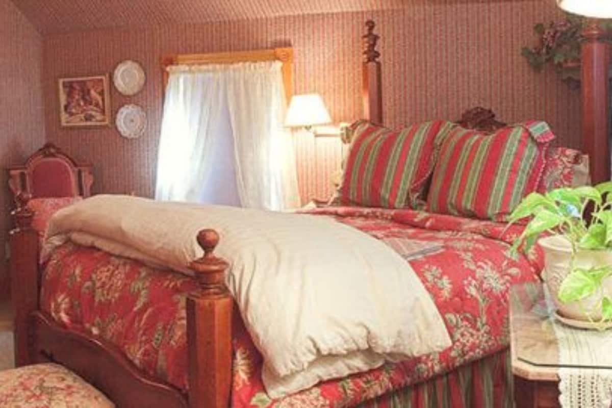 Quarto com cama de madeira, roupa de cama floral, mesa de cabeceira com abajur e um vaso com planta.