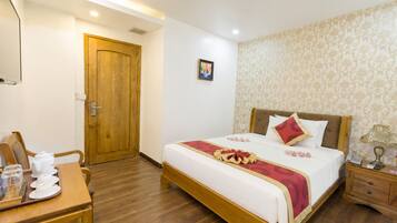 Quarto de hotel com uma cama de madeira, uma mesa de cabeceira com abajur, uma mesinha de cabeceira com telefone e uma porta.