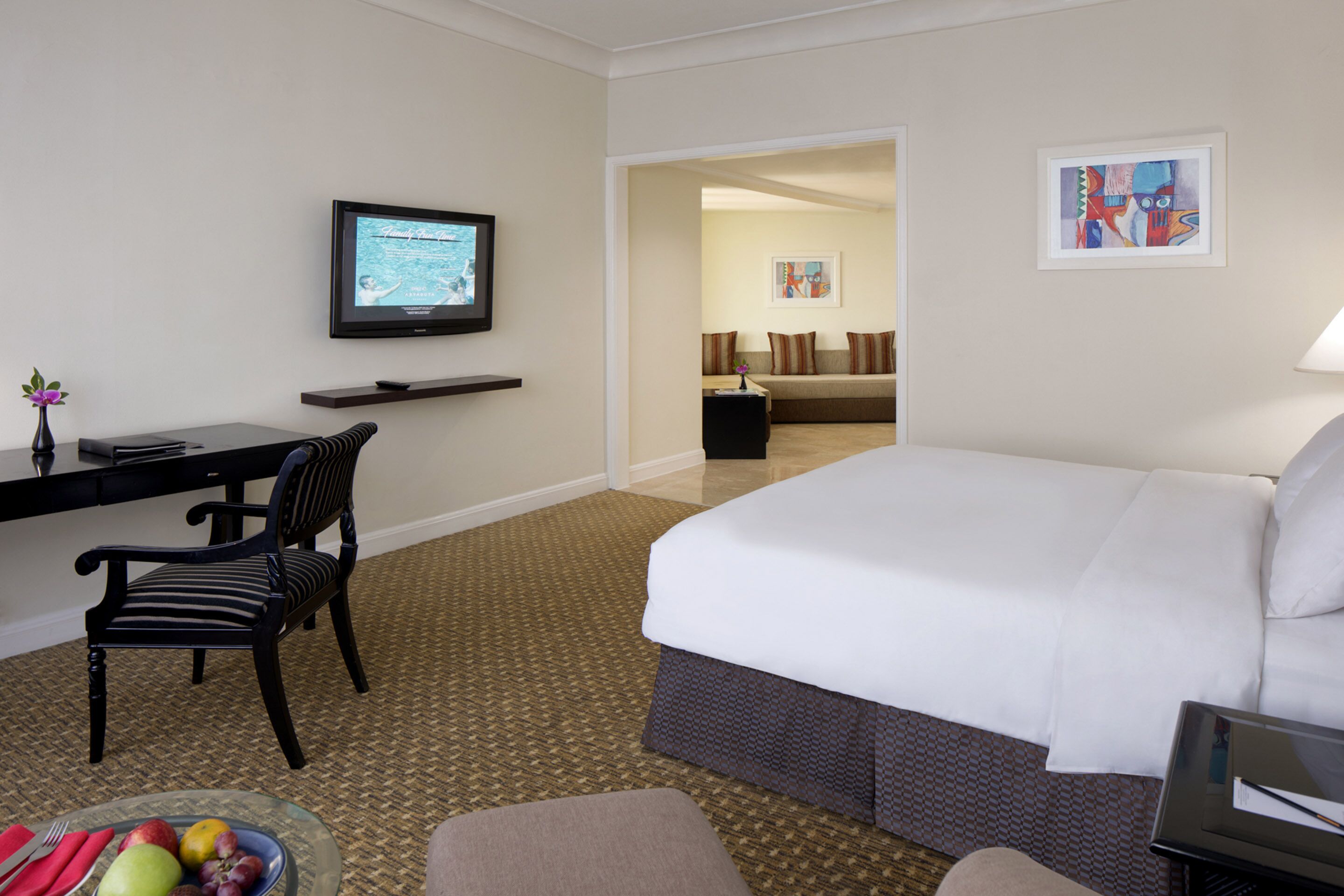 Quarto de hotel com cama, escrivaninha com computador, cadeira, pequena mesa com frutas e televisão na parede.