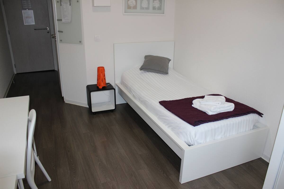 Quarto individual com uma cama de solteiro, piso de madeira, uma cama branca, uma pequena mesa com cadeira e uma porta com um aviso.