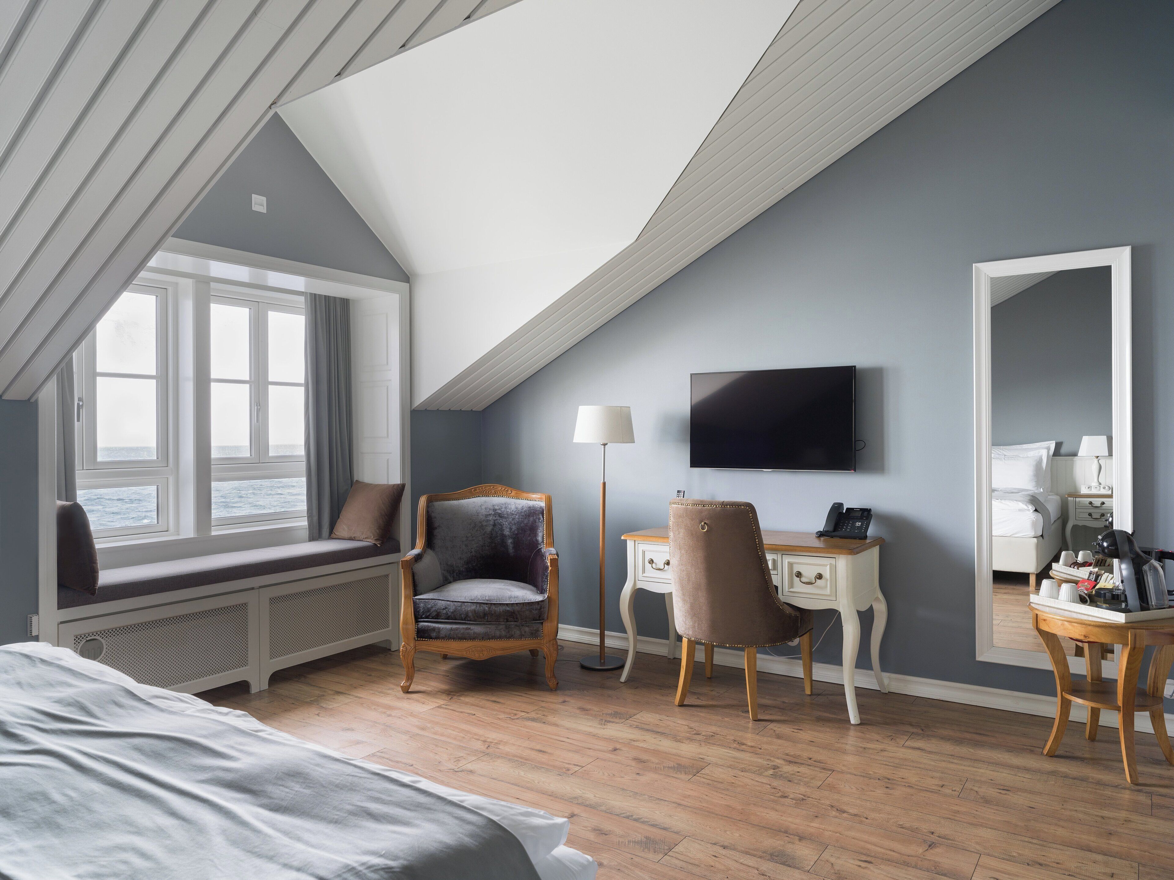 Quarto de hotel com teto inclinado, uma cama, uma mesa, uma cadeira, uma televisão e janela com vista para o mar.