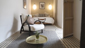 Quarto de hotel com piso xadrez, cama com travesseiros, uma cadeira, mesa pequena com um vaso e luminárias fixadas na parede.