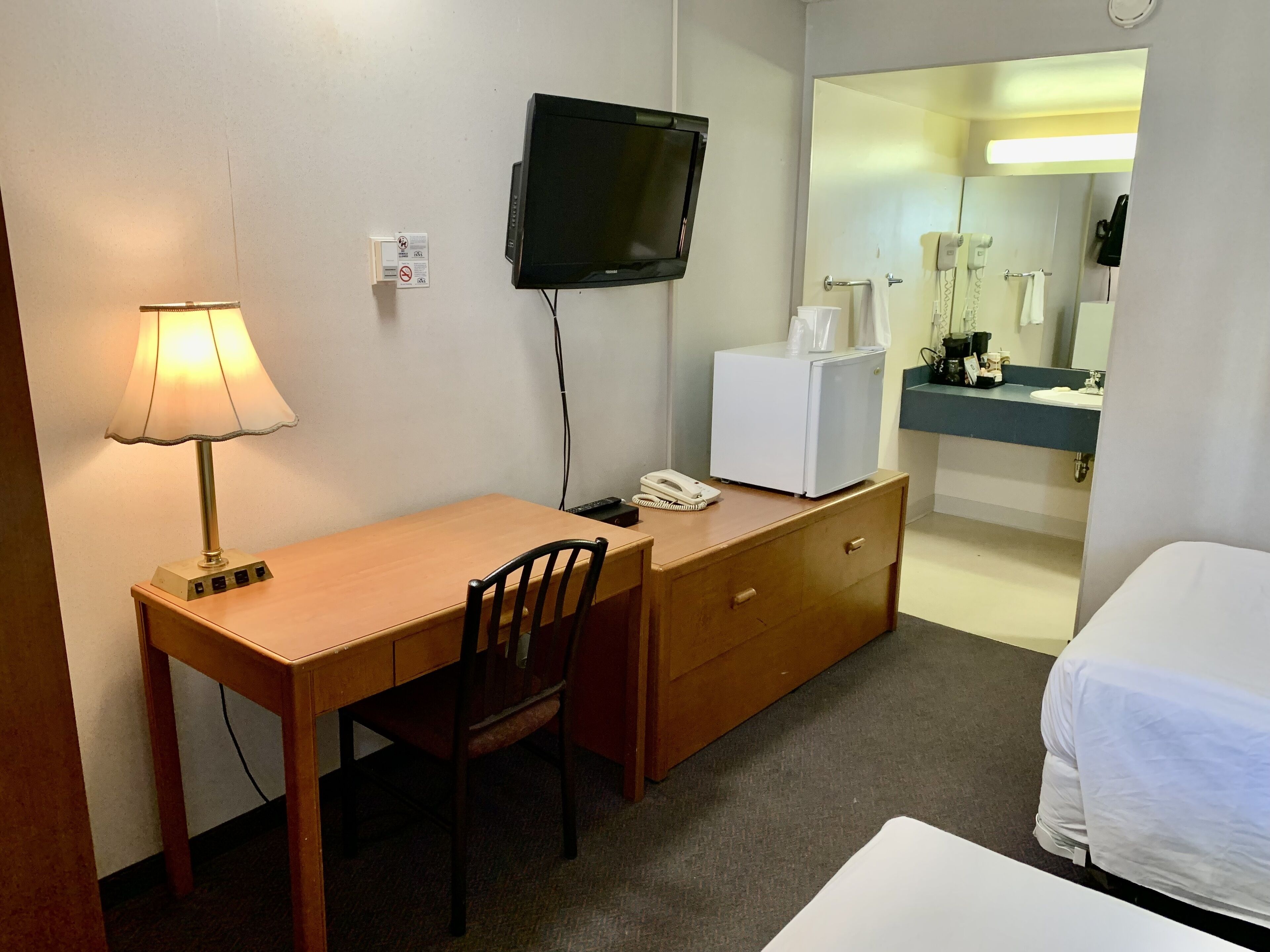 Quarto de hotel com secretária de madeira, uma cadeira, televisão presa à parede, frigorífico branco, telefone e cama com cobertura branca.