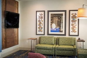 Quarto de hotel com um sofá verde, uma TV de tela plana fixada numa parede de madeira e três obras de arte emolduradas acima do sofá.