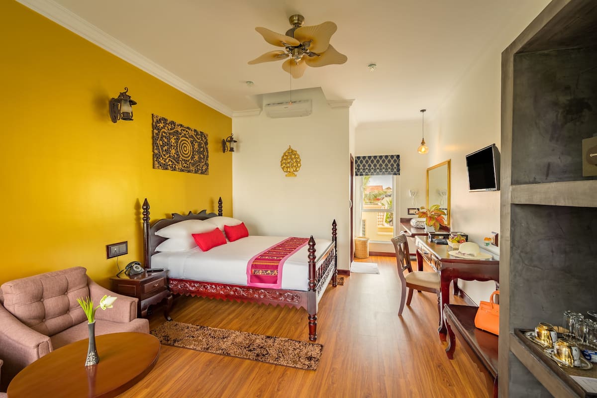 Quarto de hotel com cama, escrivaninha, cadeira, mesinha com um vaso e ventoinha no teto.