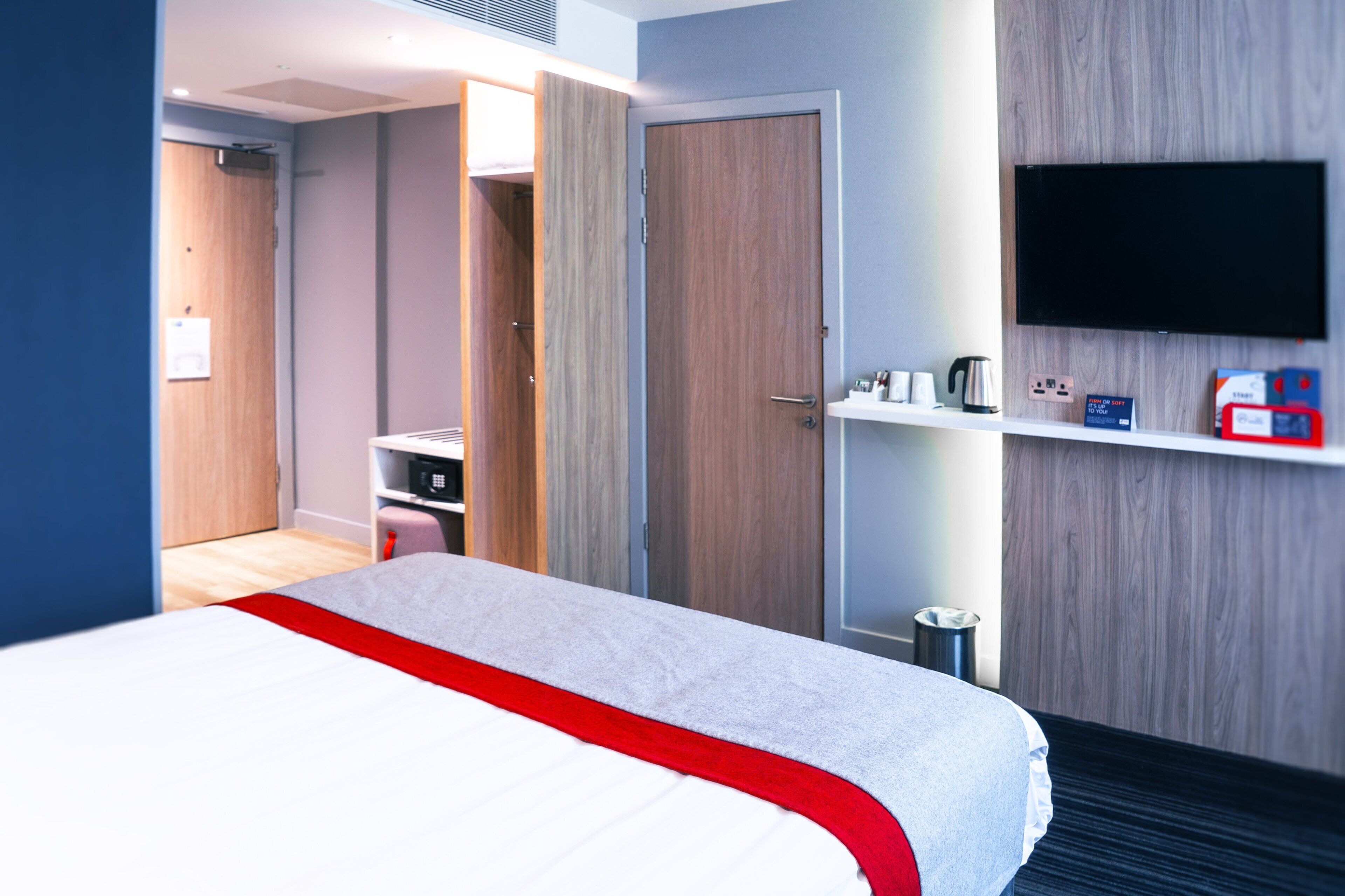 Quarto de hotel com uma cama, uma televisão fixada na parede, um pequeno armário com produtos de higiene e uma porta para o banheiro.