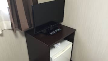 Quarto de hotel compacto com televisão de tela plana em escrivaninha de madeira escura, pequena geladeira branca embaixo e parede lisa com textura.