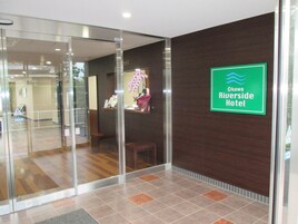 Área de recepção de hotel com entrada por porta de vidro, balcão de recepção e placa indicativa do Hotel Okawa Riverside.
