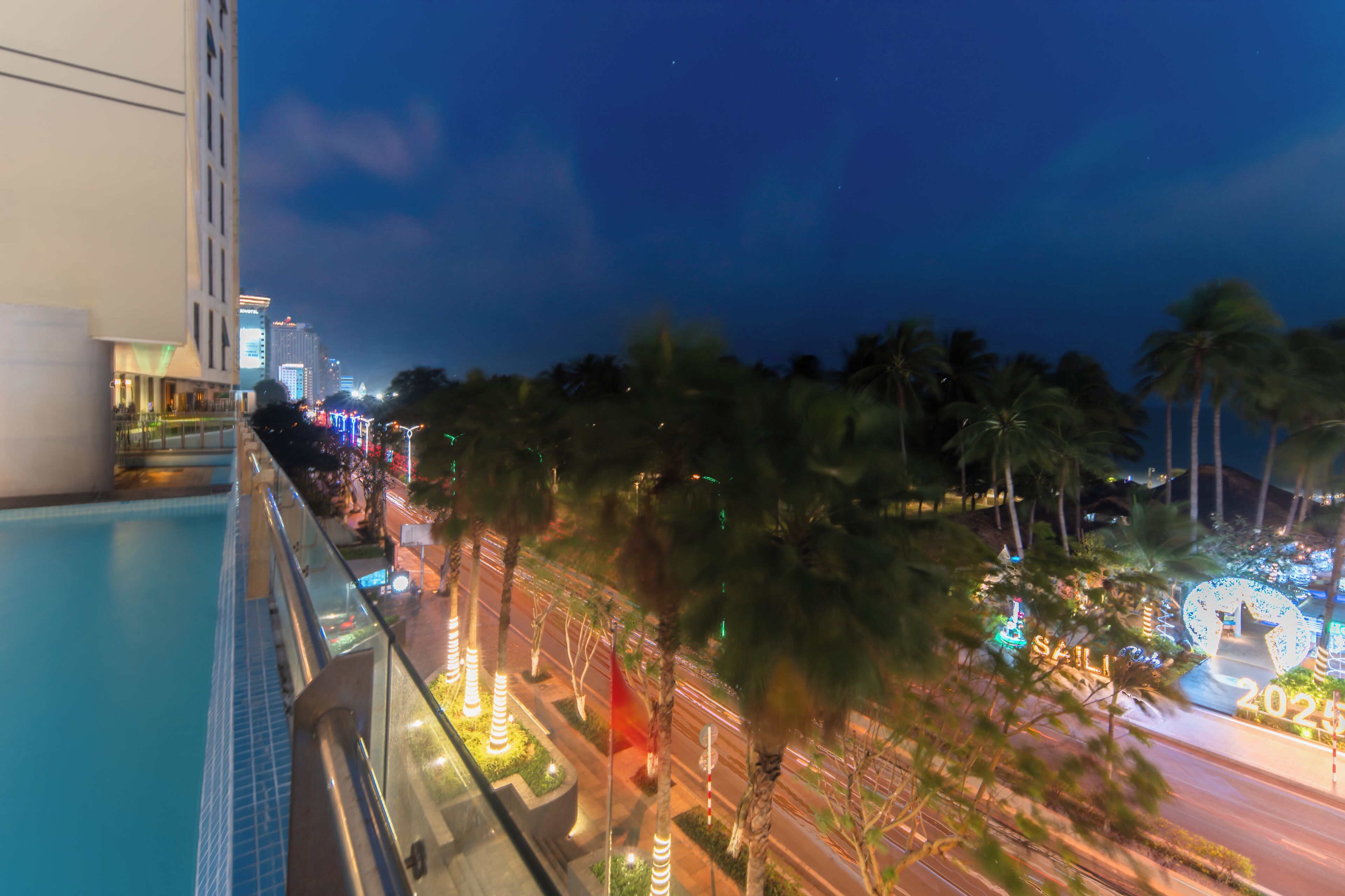 Vista noturna de um hotel com piscina, palmeiras e postes de luz iluminados.