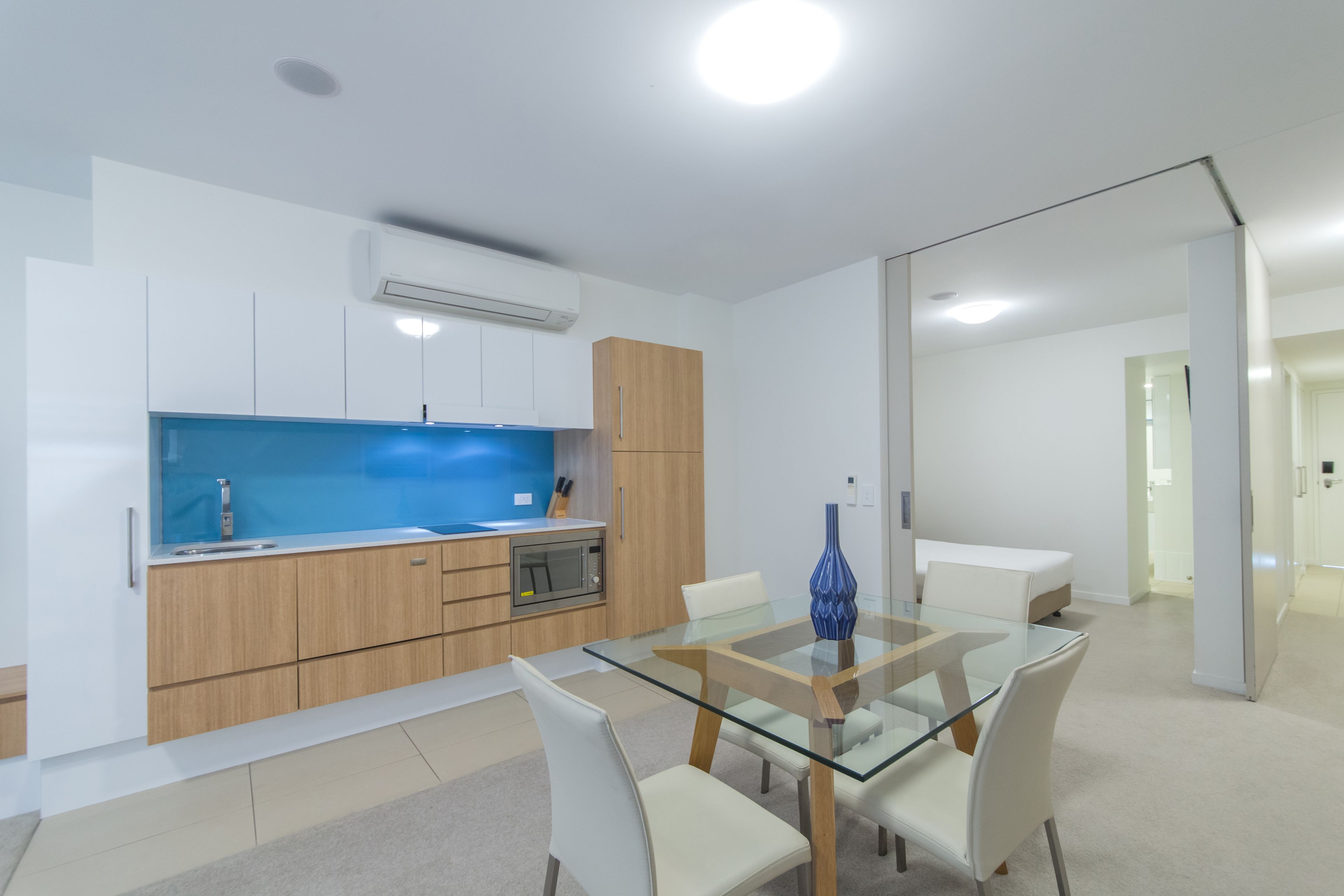 Uma cozinha moderna com armários brancos, painel de fundo azul e uma mesa de jantar de vidro com cadeiras brancas.