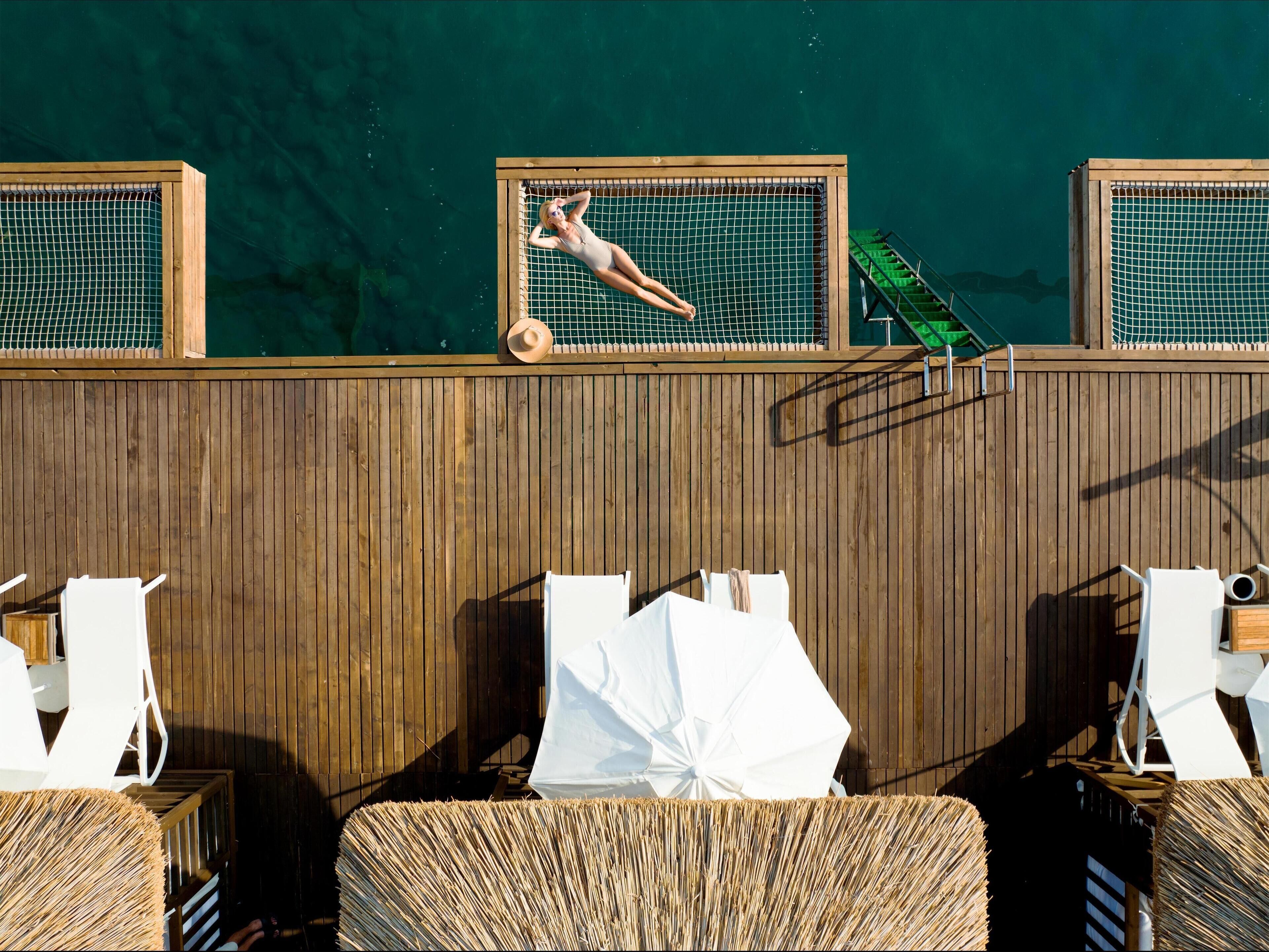 Um deck de madeira com espreguiçadeiras e guarda-sóis, com vista para uma área de piscina com trampolim e um chapéu no corrimão.