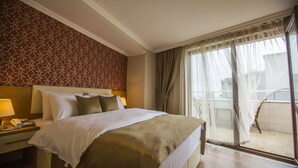 Quarto de hotel com uma cama grande, uma mesinha de cabeceira com abajur, papel de parede estampado e uma varanda com vista.