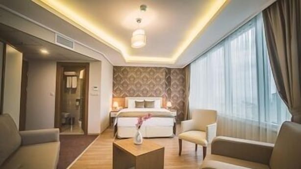Quarto de hotel com cama, duas poltronas, uma mesa de centro pequena e uma janela grande com cortinas.