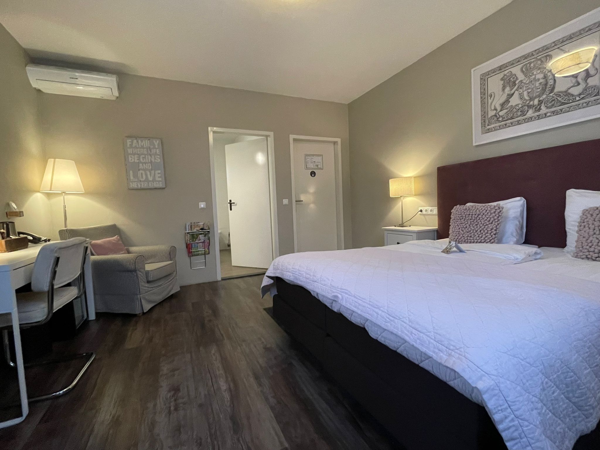 Quarto de hotel com cama, escrivaninha, cadeira, abajur e porta.