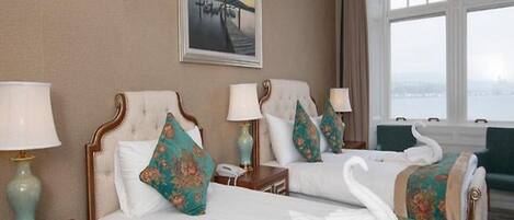 Quarto de hotel com duas camas, cada uma com colcha branca e cabeceiras combinando, ladeadas por mesas de cabeceira com abajures. Uma moldura com um quadro está pendurada acima das camas, e uma janela com cortinas oferece vista para o exterior.