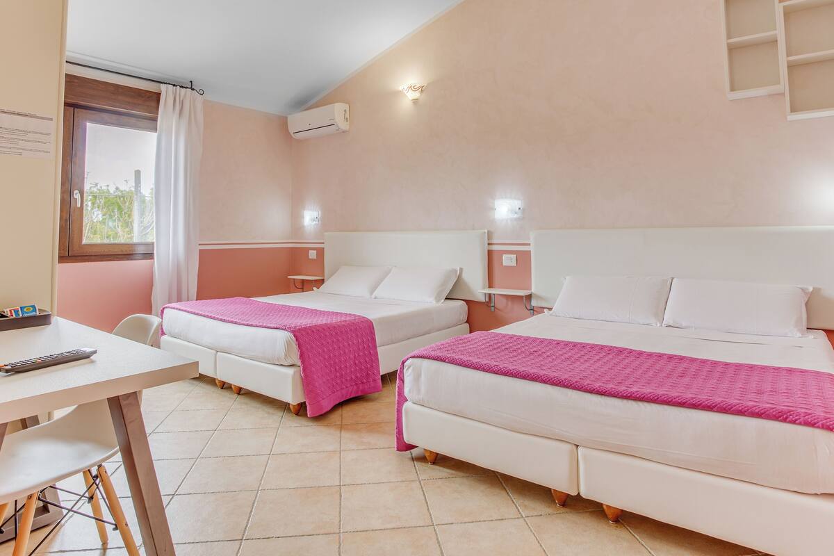 Quarto de hotel com duas camas, uma escrivaninha, janela com cortinas, ar condicionado e prateleira fixada na parede.