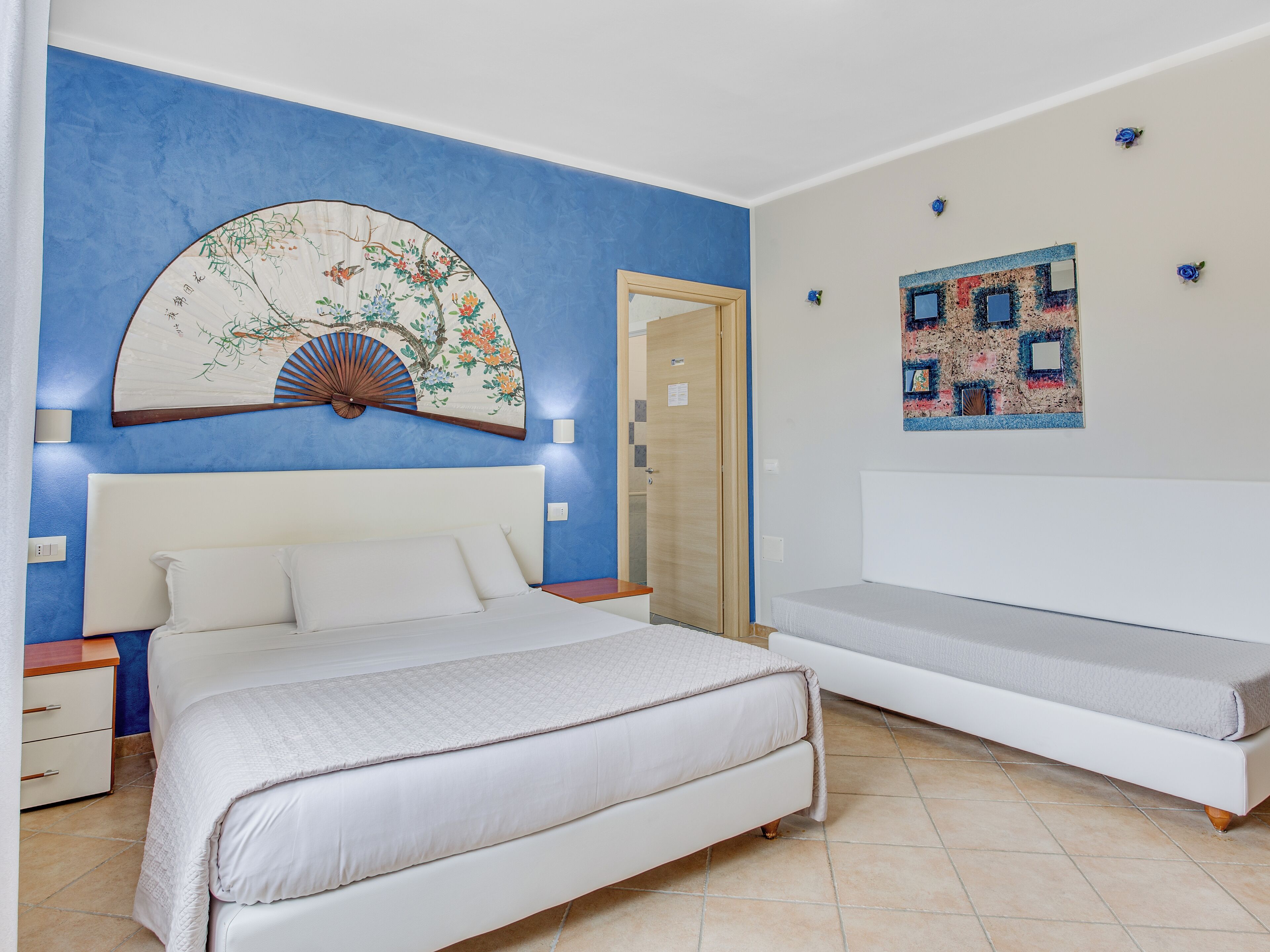 Quarto com cama, um banco, um ventilador e parede com pintura floral.