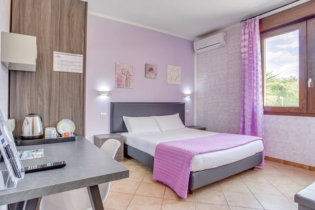Quarto de hotel com cama, mesa de cabeceira e janela com cortinas.