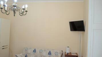 Quarto com parede bege, porta branca, armário de madeira, sofá com padrão náutico, televisão na parede e um lustre.
