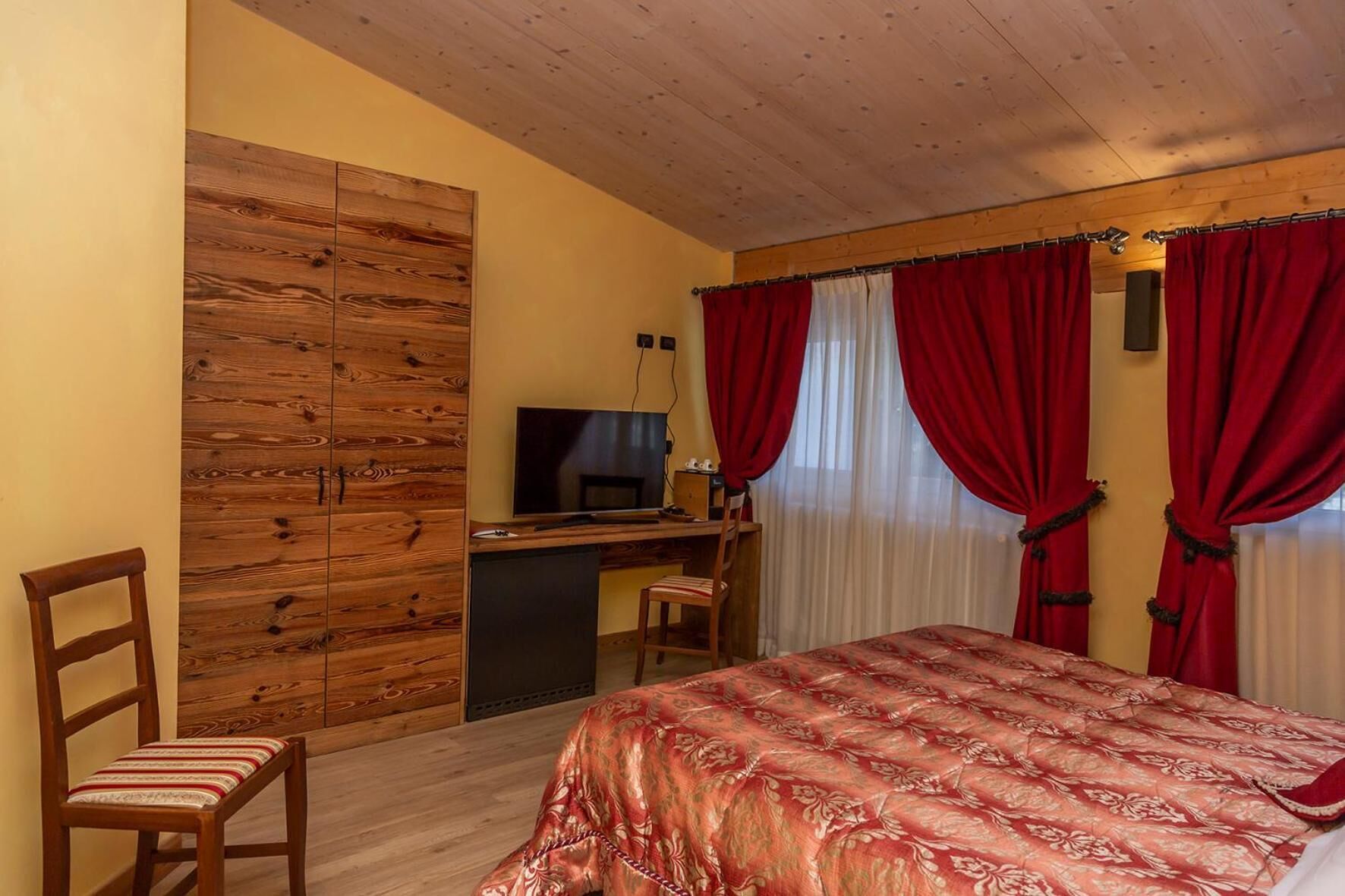 Quarto com guarda-roupa de madeira, escrivaninha com televisão, cama com colcha vermelha e dourada, e cortinas vermelhas.