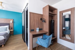 Quarto de hotel pequeno com cama, escrivaninha, cadeira, espelho e guarda-roupa.