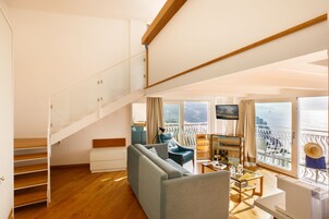 Uma sala de estar ampla com sofá, poltronas, mesa de centro e televisão. Uma escada leva a um andar superior.