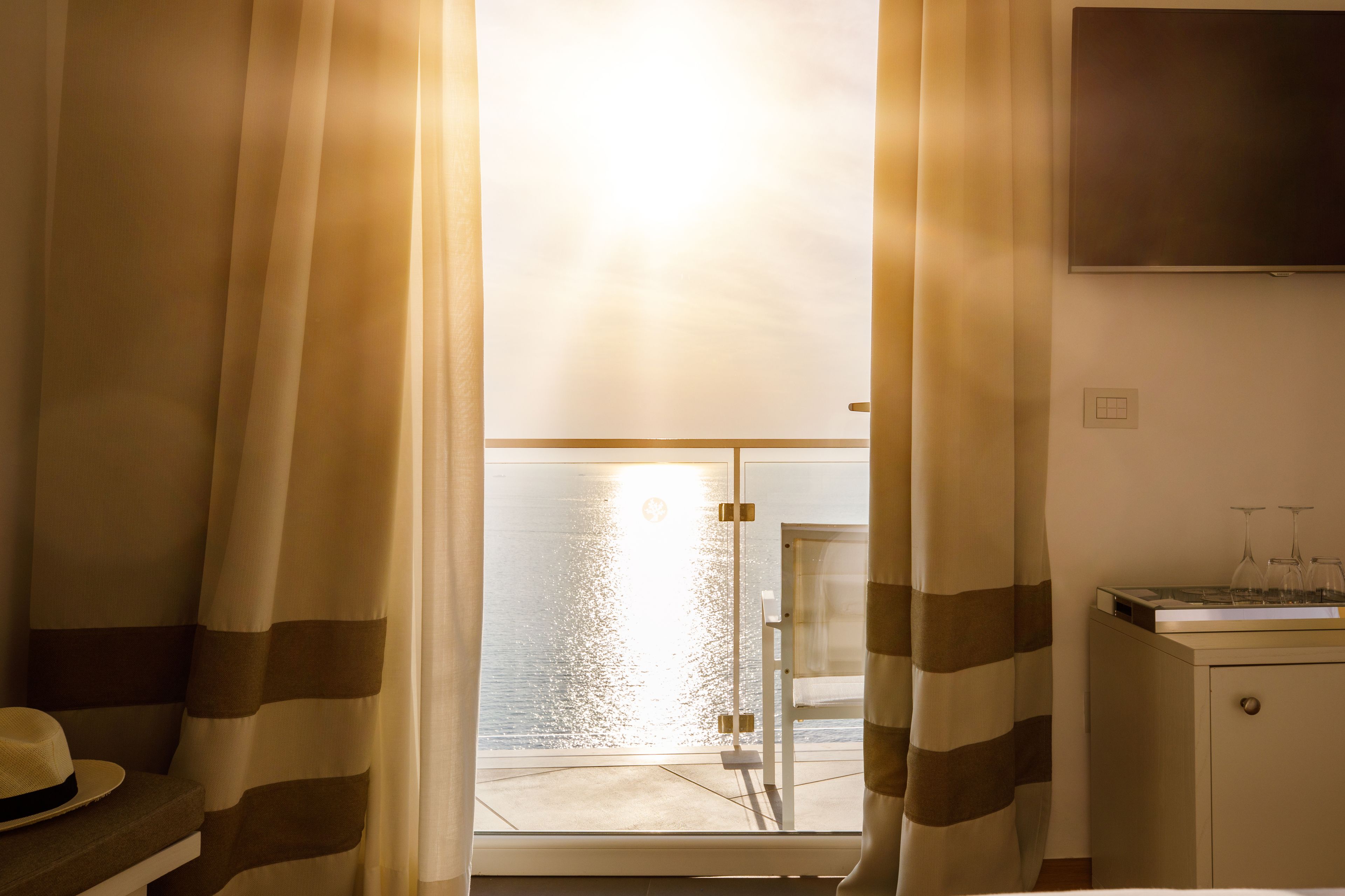 Quarto de hotel com vista para o mar através de uma porta de vidro, mesa de cabeceira com chapéu e televisão presa à parede.