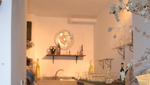 Uma cozinha com um vaso branco em uma mesa, uma estante com garrafas e uma planta pendente.