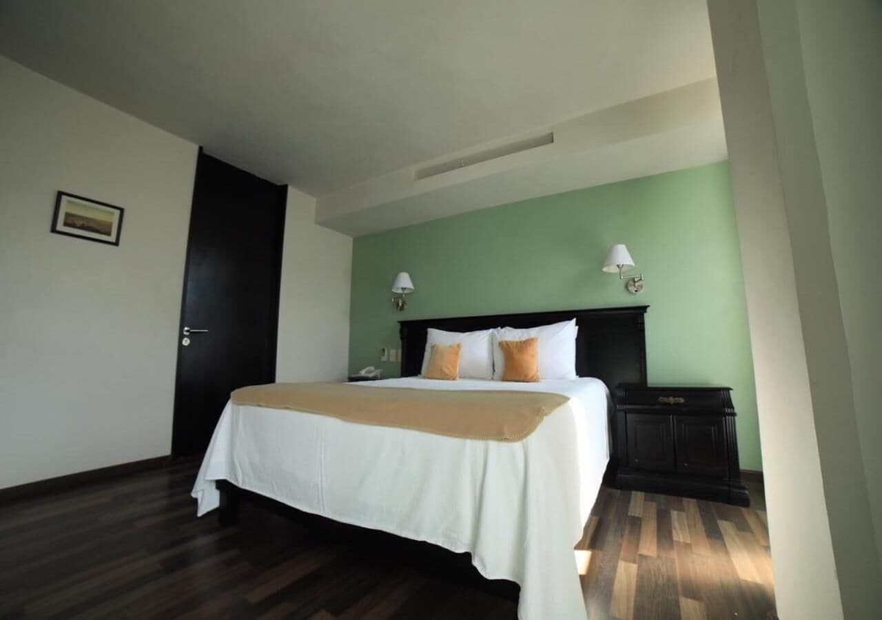 Cama bem arrumada com colcha branca e bege, cabeceira de madeira escura e duas luminárias de cabeceira.