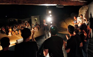 Um palco com um orador e plateia sentada no chão.