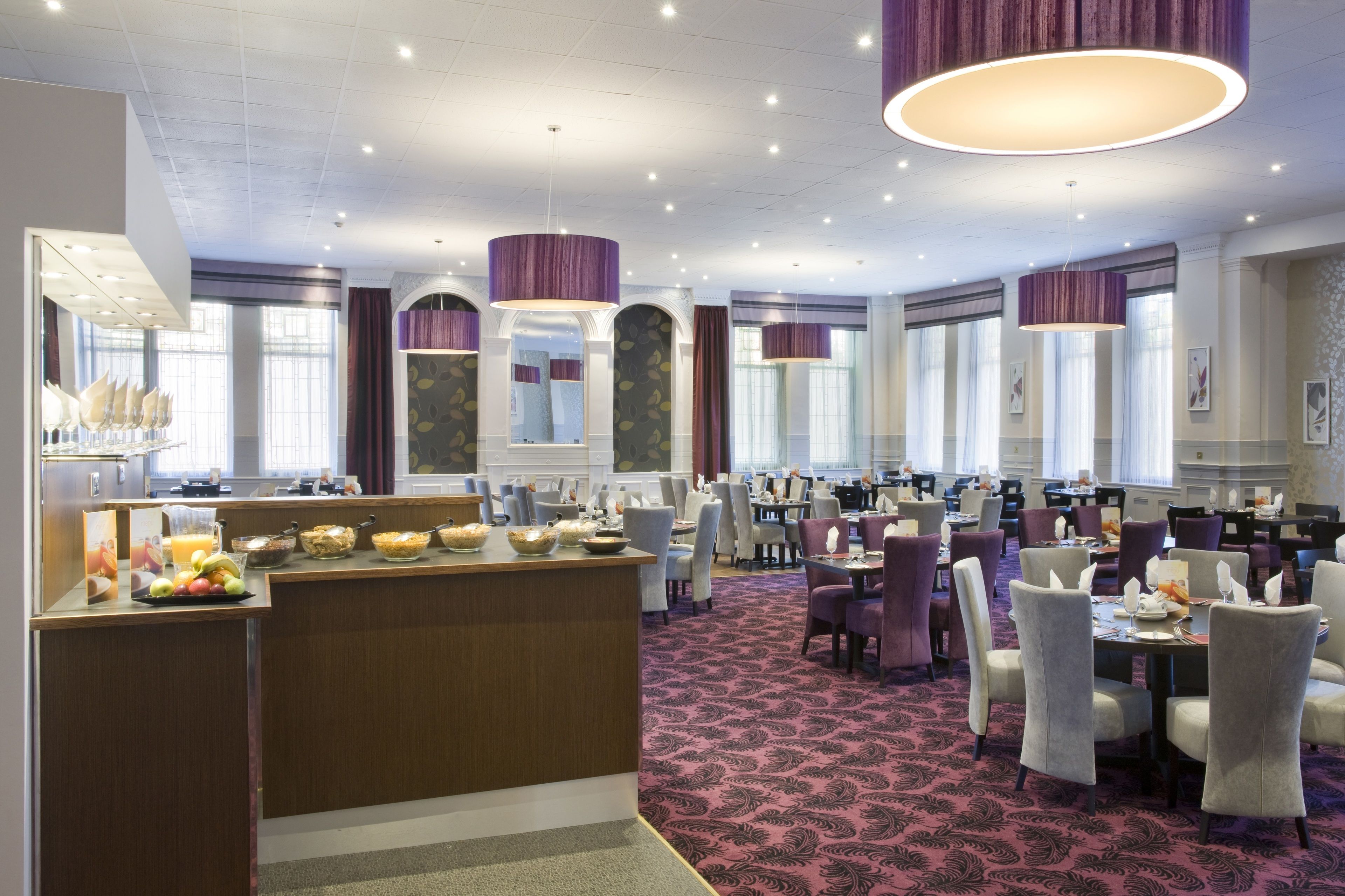Uma sala de jantar de hotel com várias mesas e cadeiras, um balcão de recepção e um tapete estampado.