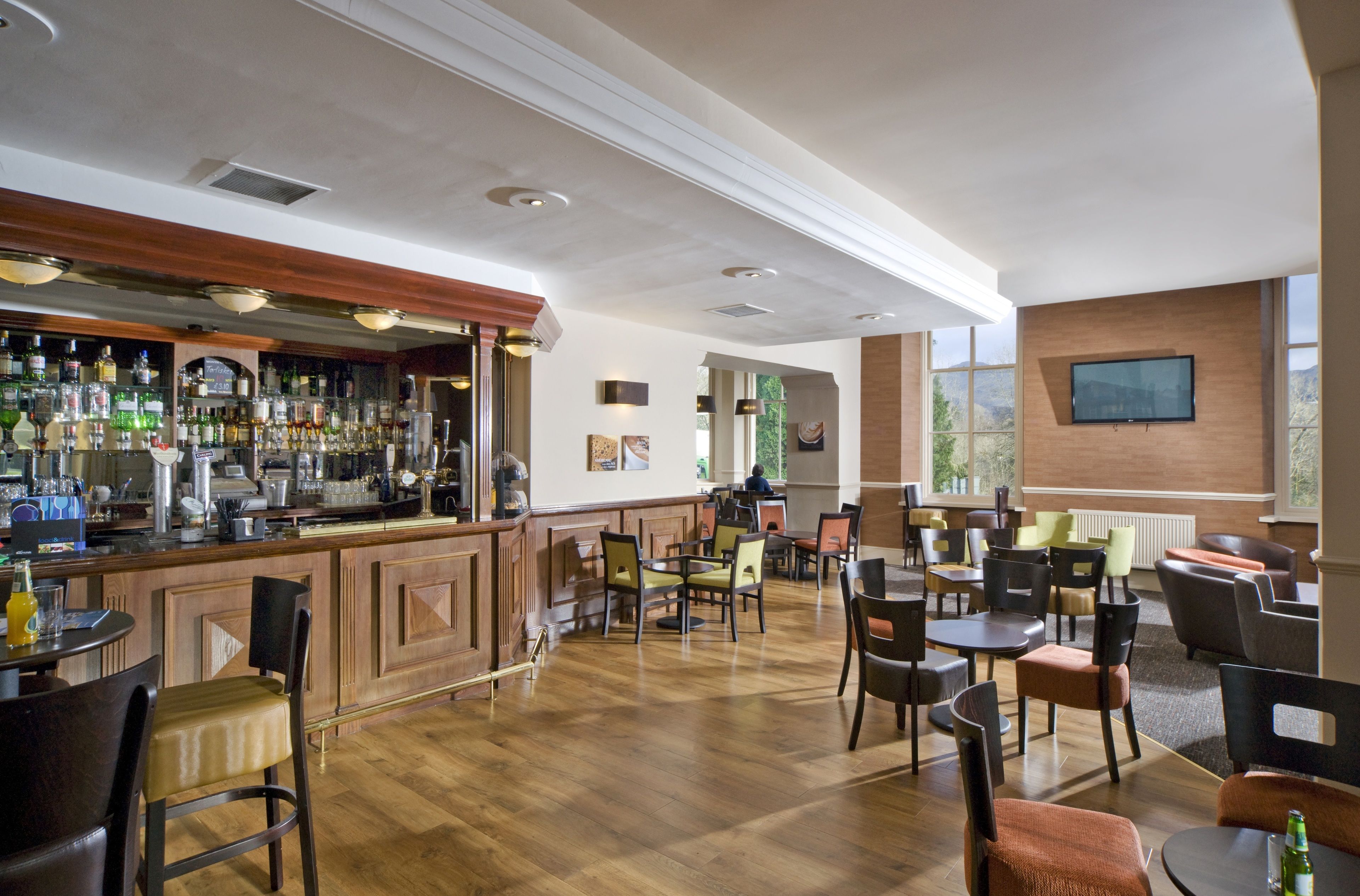 Uma área de bar com balcão de madeira e diversos tipos de bebidas, rodeada por várias opções de assentos, incluindo cadeiras e mesas.