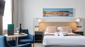 Quarto de hotel com uma cama, duas mesas de cabeceira, uma cadeira, uma mesinha com um menu e uma garrafa, e um quadro de uma cidade costeira na parede.