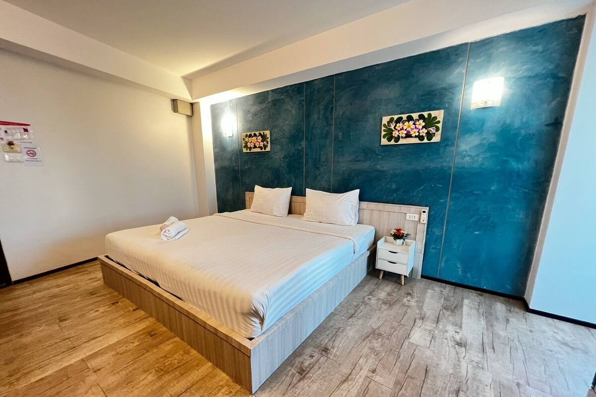 Quarto de hotel moderno com uma cama grande, piso de madeira, parede azul e uma pequena mesa de cabeceira com um vaso de flores.