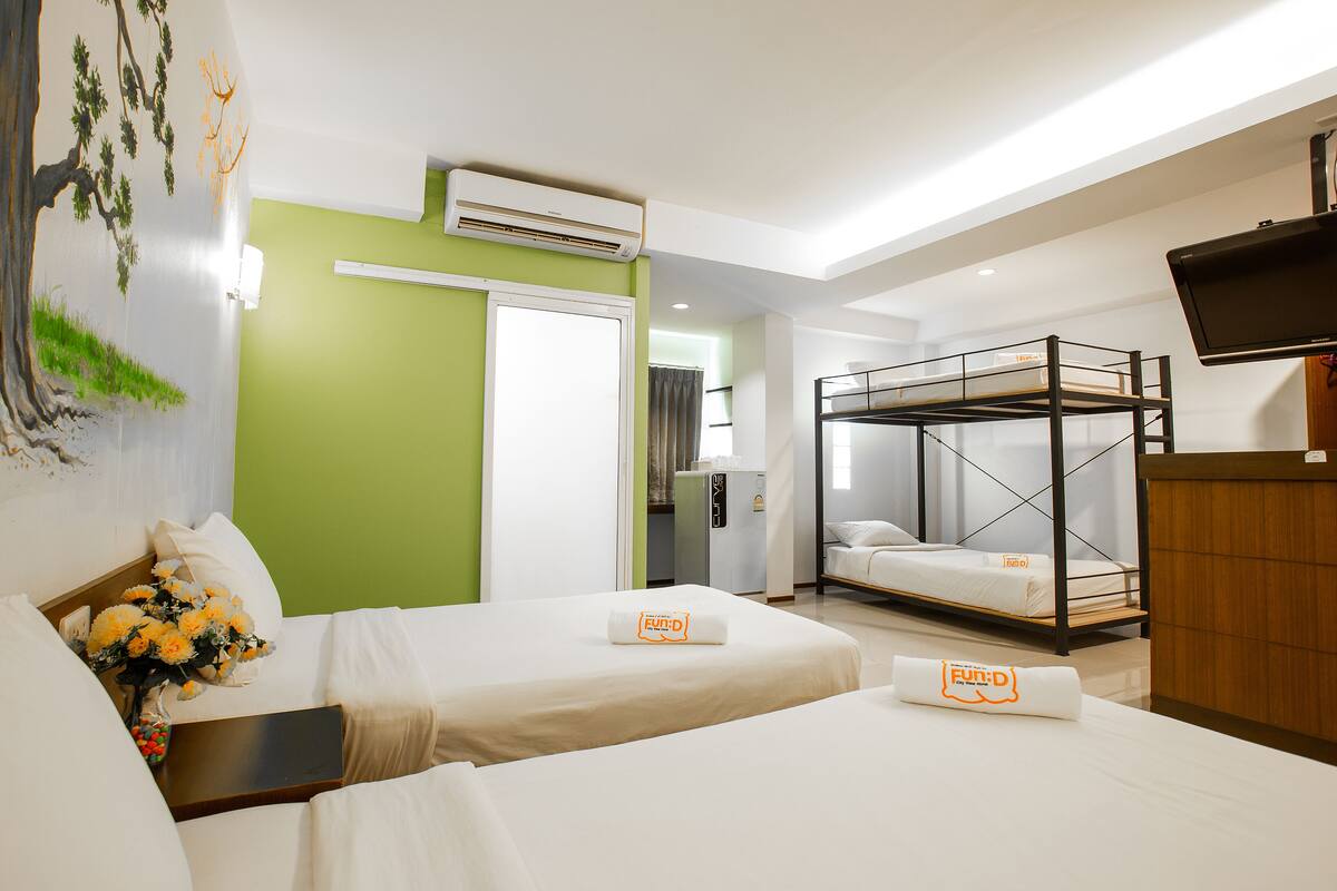 Quarto de hotel com duas camas, uma cama beliche, televisão de tela plana, ar condicionado e uma parede verde.