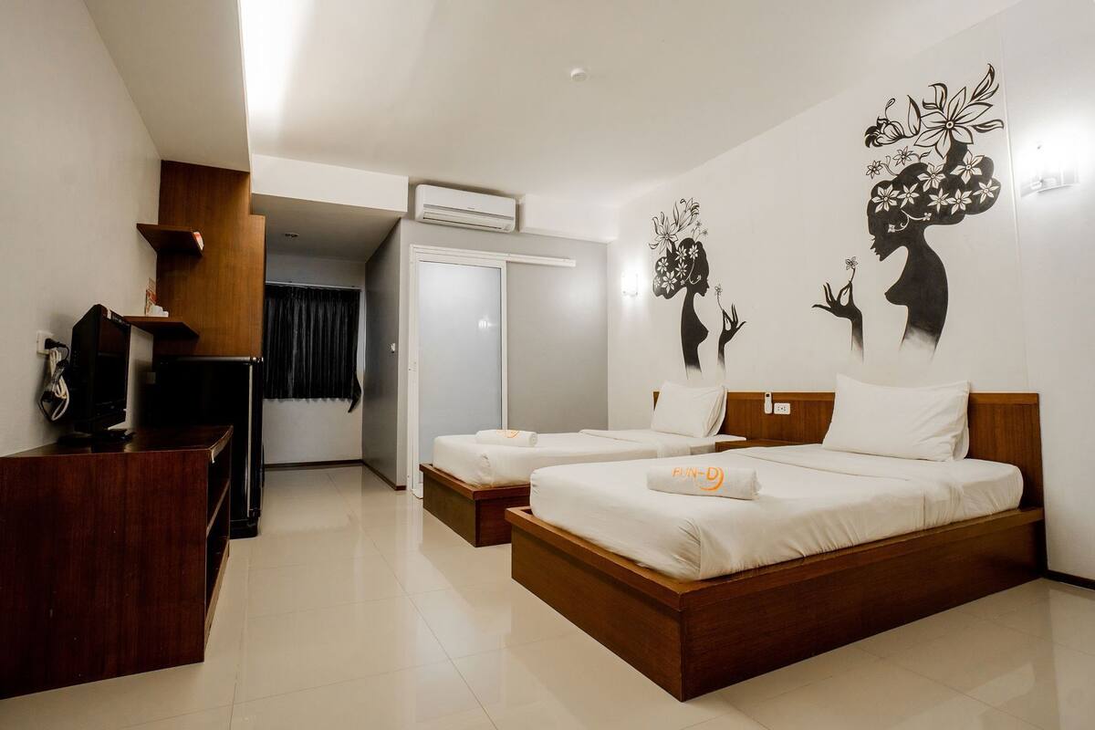 Quarto de hotel com duas camas, televisão, guarda-roupa e parede com arte floral.